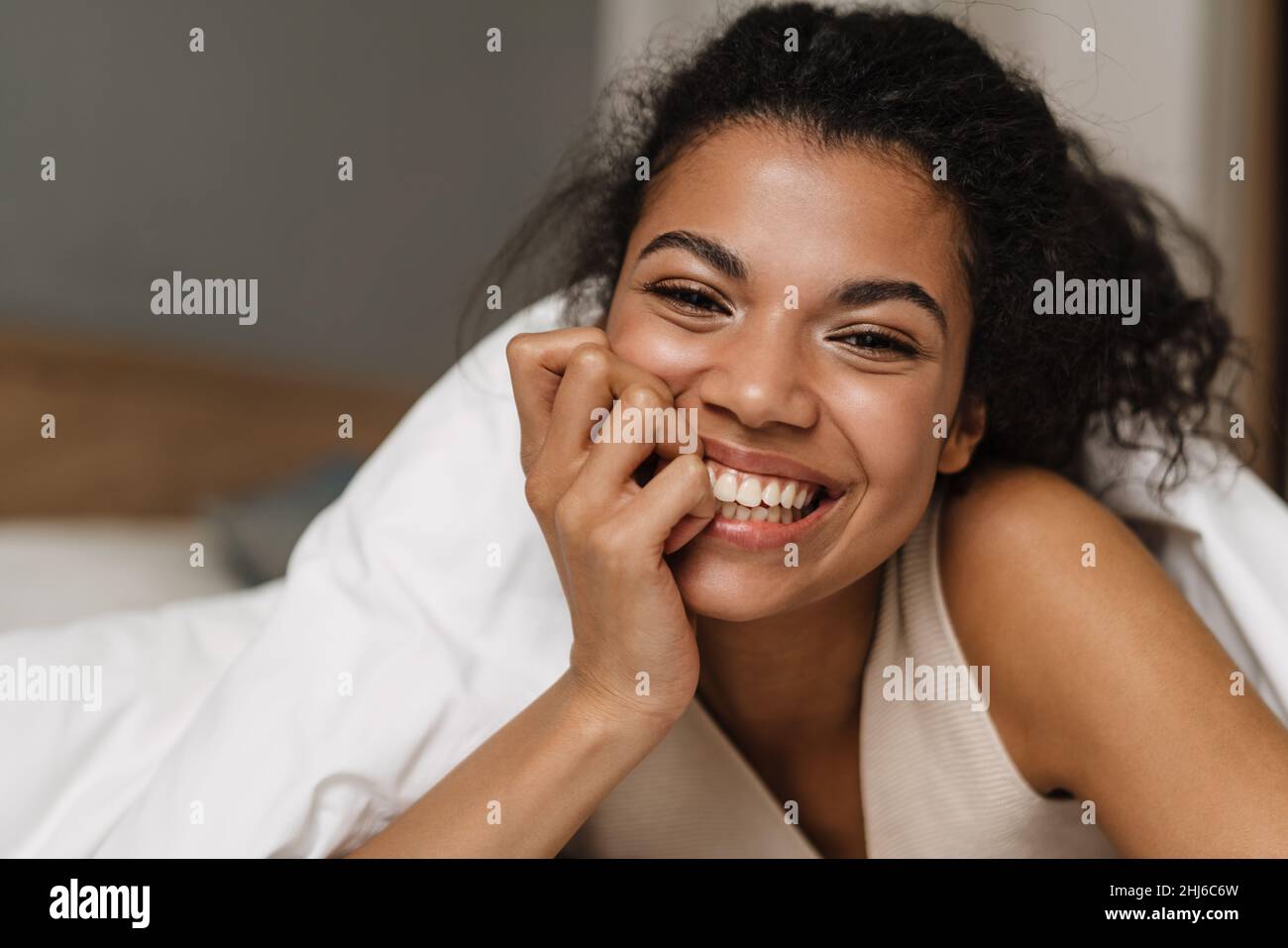 Happy young african woman laying hiding under a blanket on a bed at ...