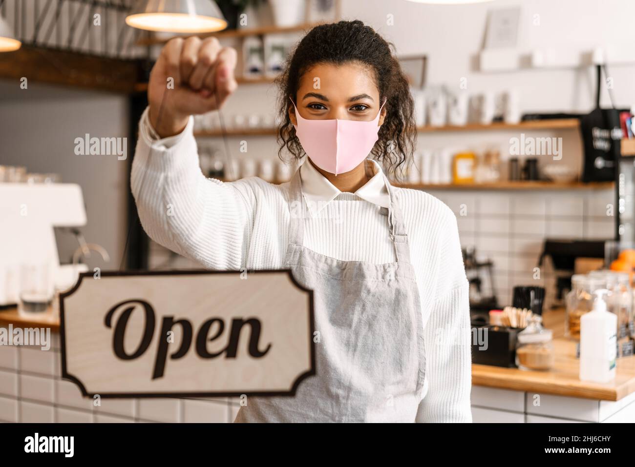 Young black waitress in face mask showing open sign board at cafe ...