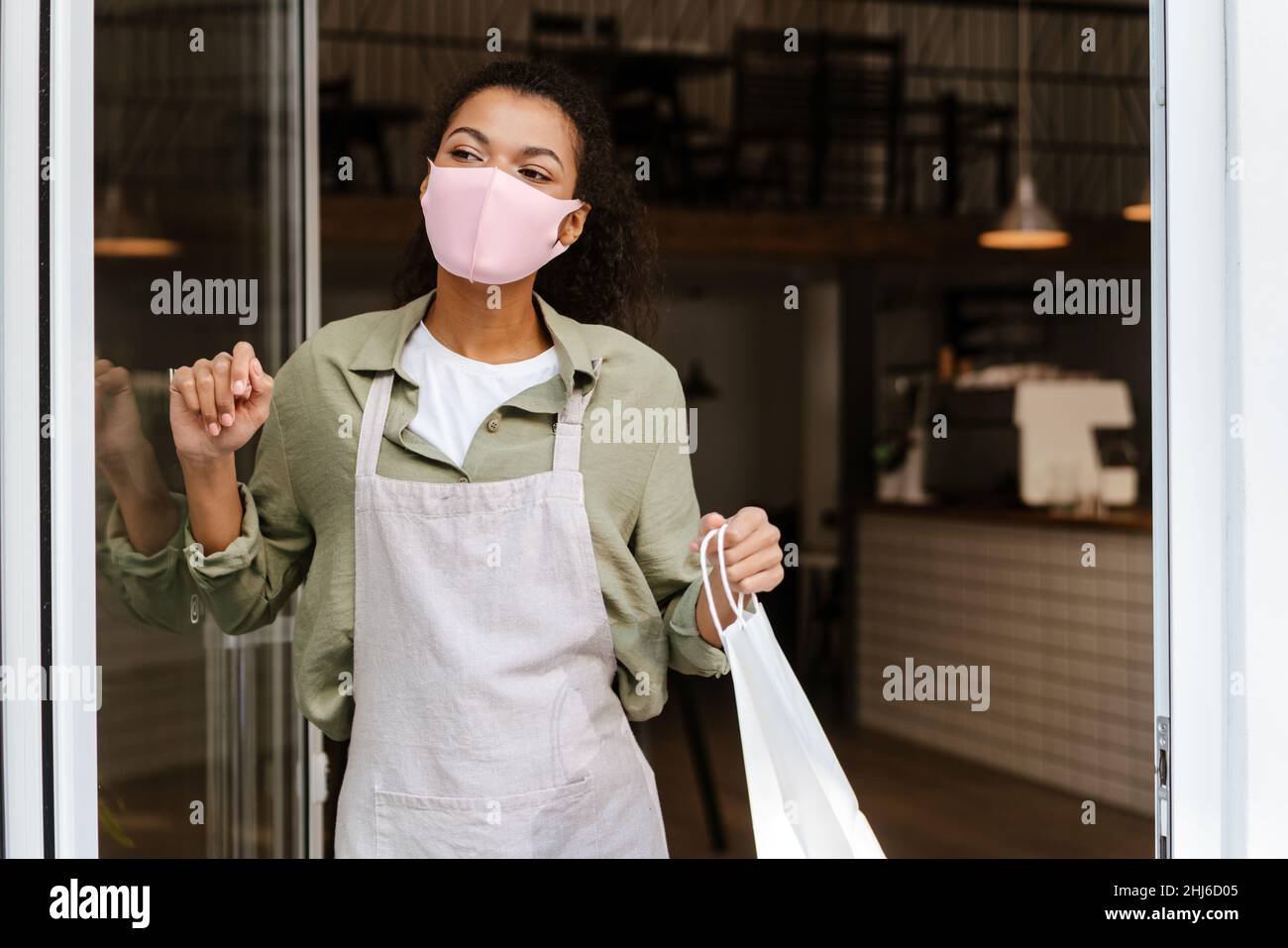 Black waitress wearing face mask holding paper bag while standing in ...