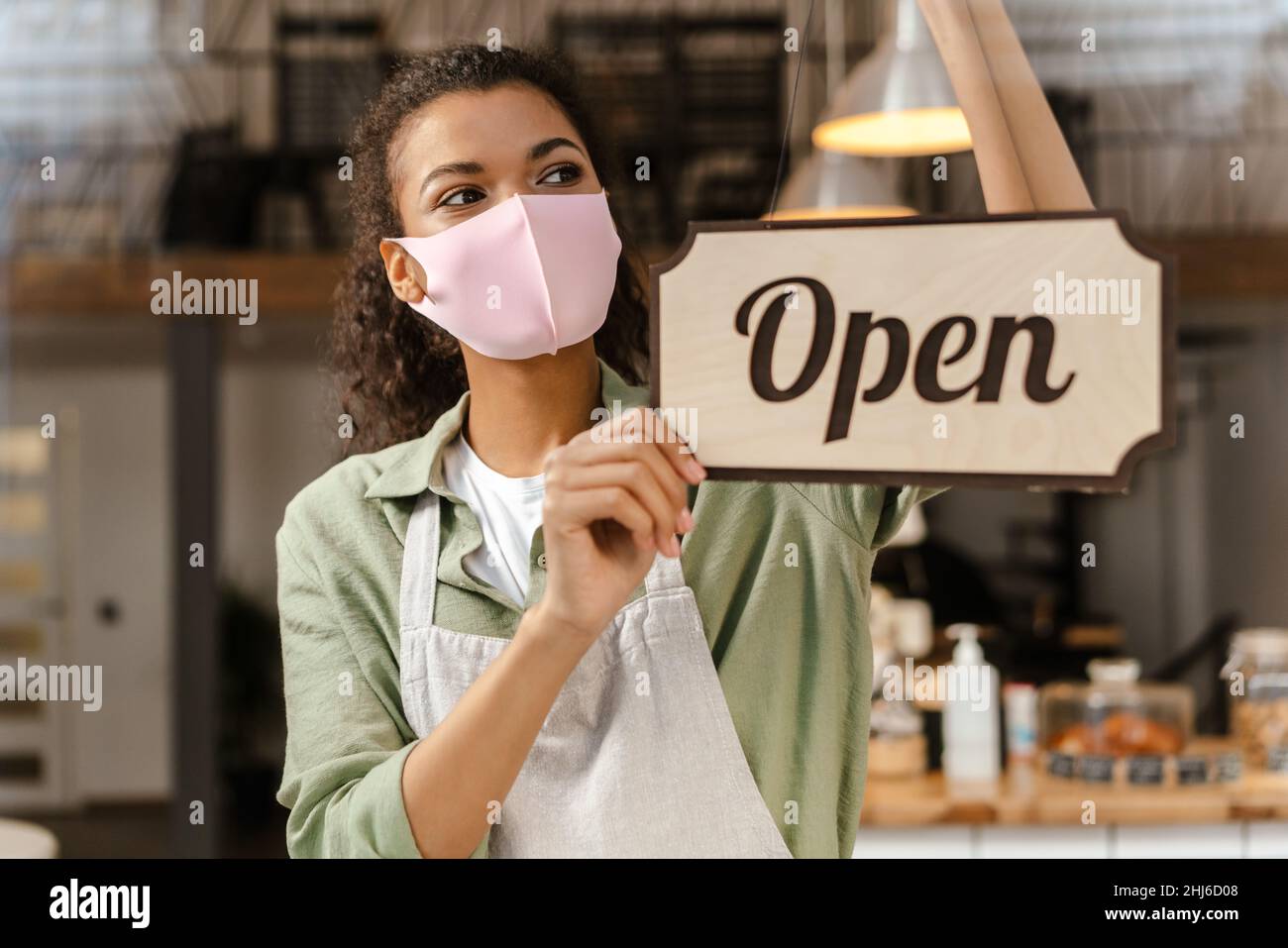 Young black waitress in face mask showing open sign board at cafe ...