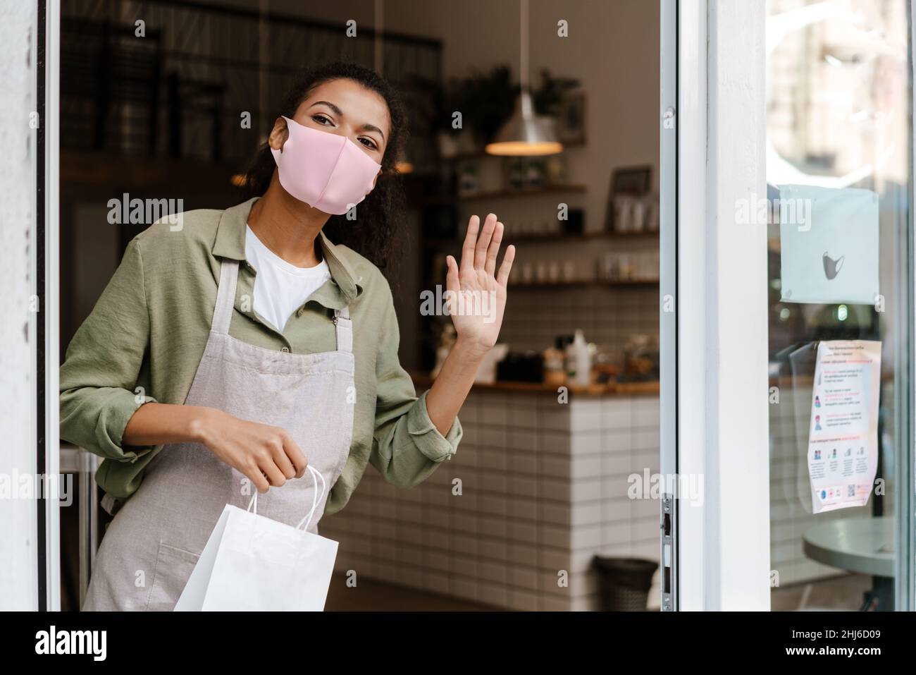 Black waitress wearing face mask holding paper bag while standing in ...