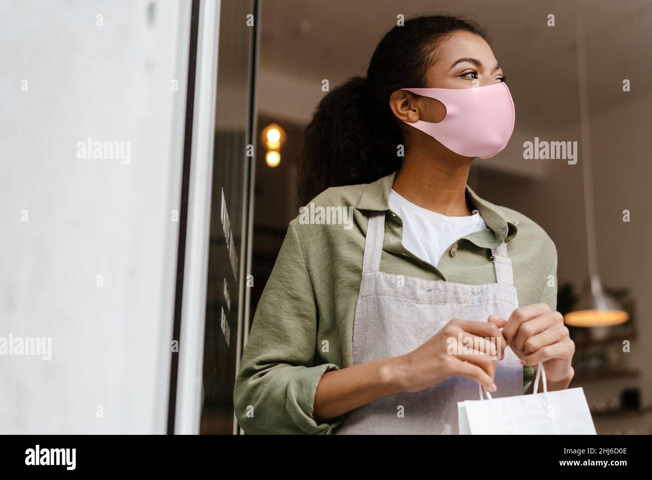 Black waitress wearing face mask holding paper bag while standing in ...