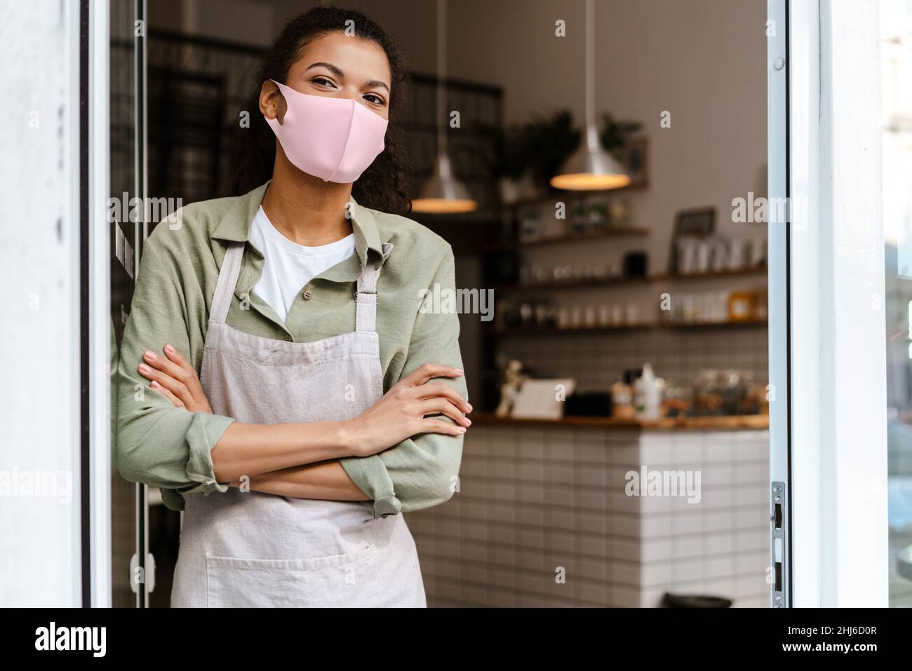 Young black waitress wearing face mask standing in doorway at cafe ...
