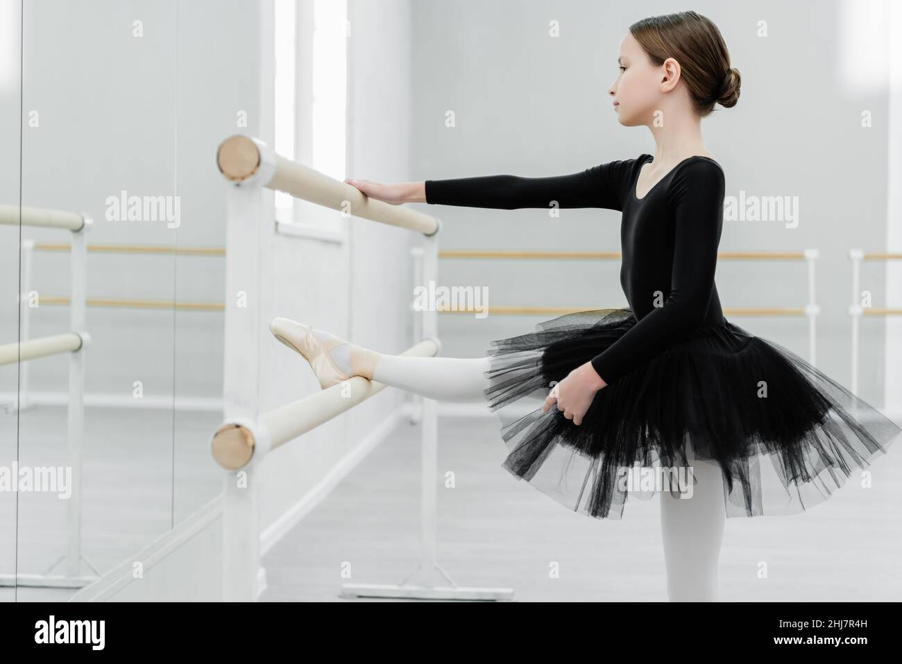 side view of kid in black tutu stretching at barre in dancing hall ...