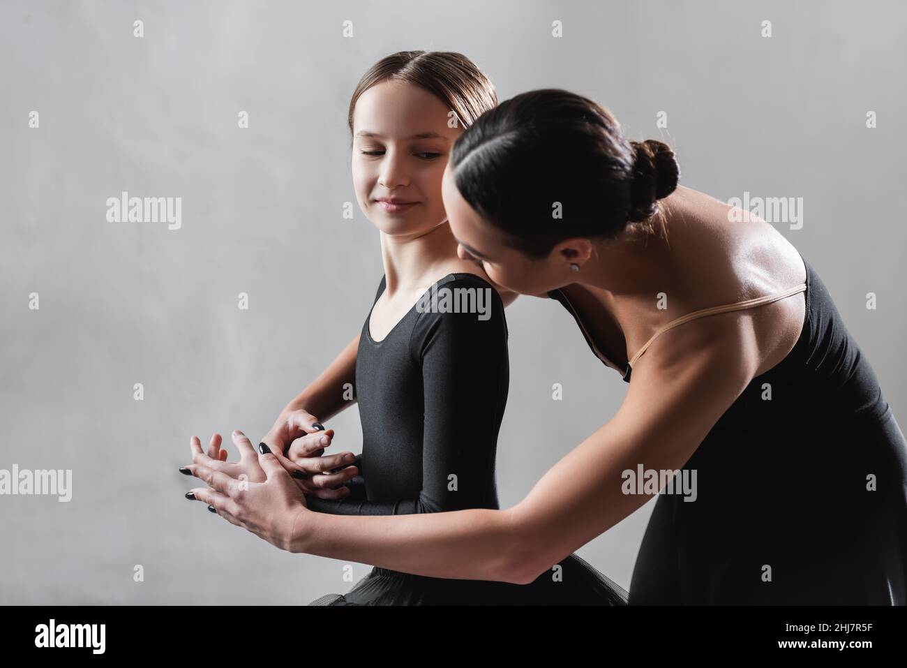 young ballet teacher touching hands of girl during dance lesson on grey ...