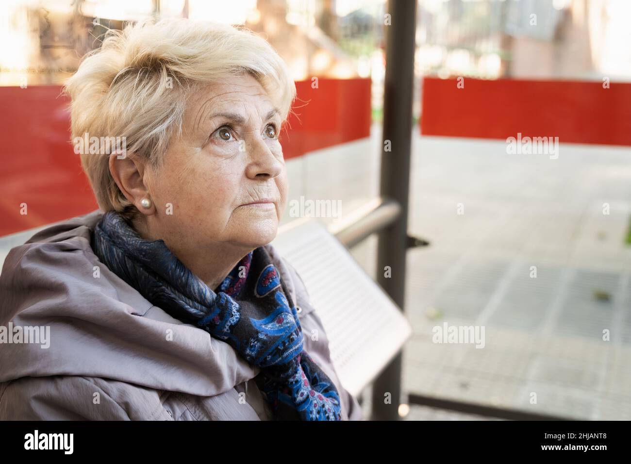Portrait of senior blonde woman sitting on bus stop. Serious elder lady ...