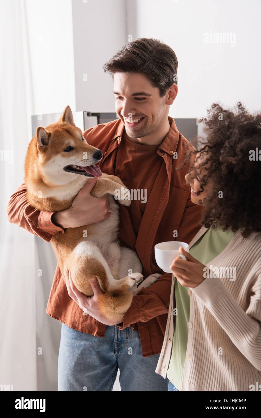 joyful man holding shiba inu dog near african american girlfriend with ...