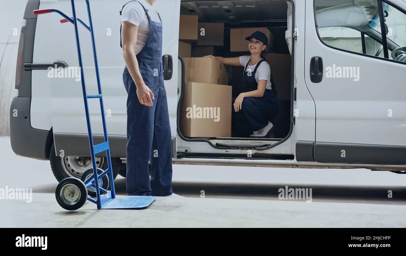 Smiling delivery woman working with carton boxes and colleague near car ...