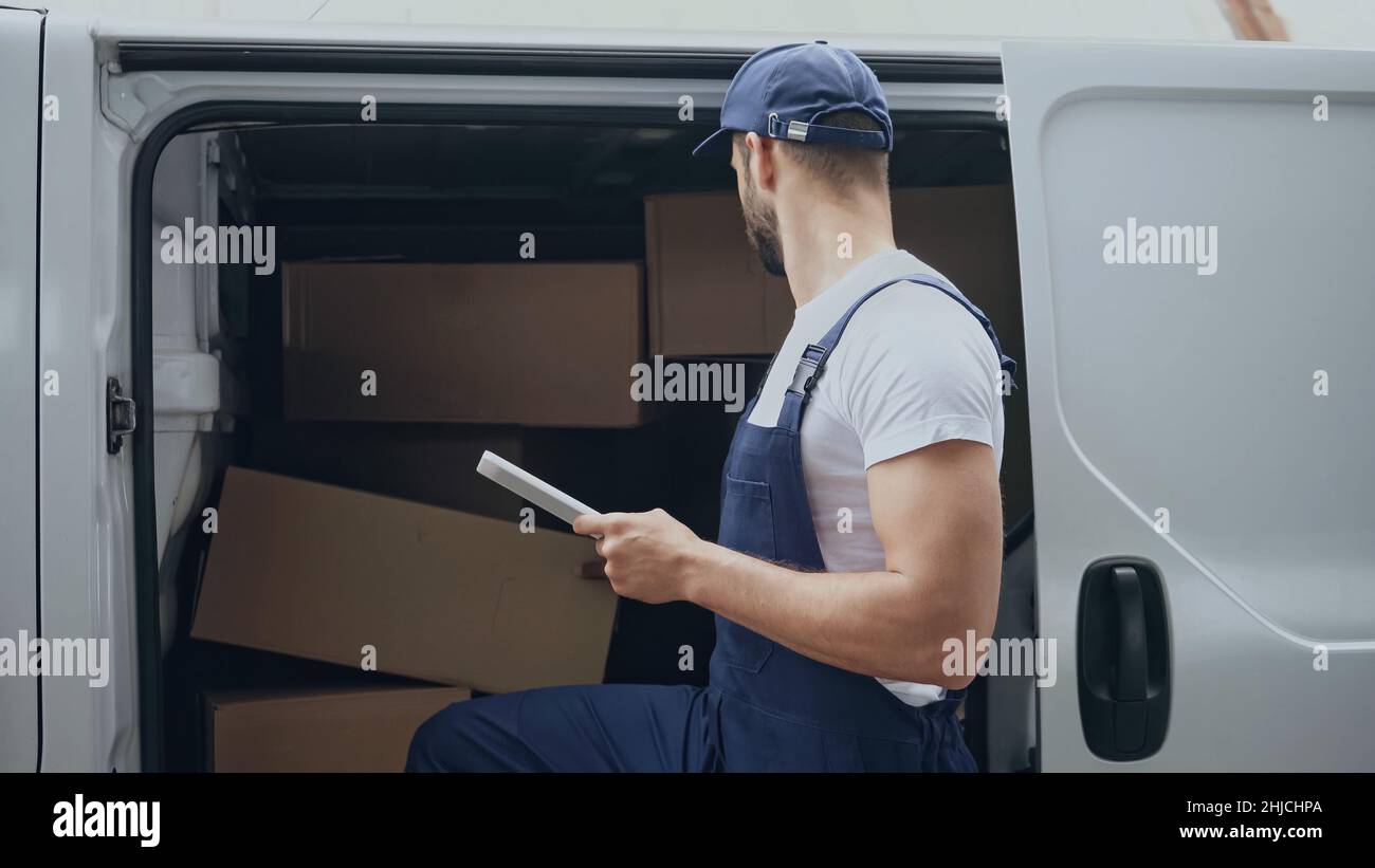 Delivery man in overalls holding digital tablet near carton boxes in ...