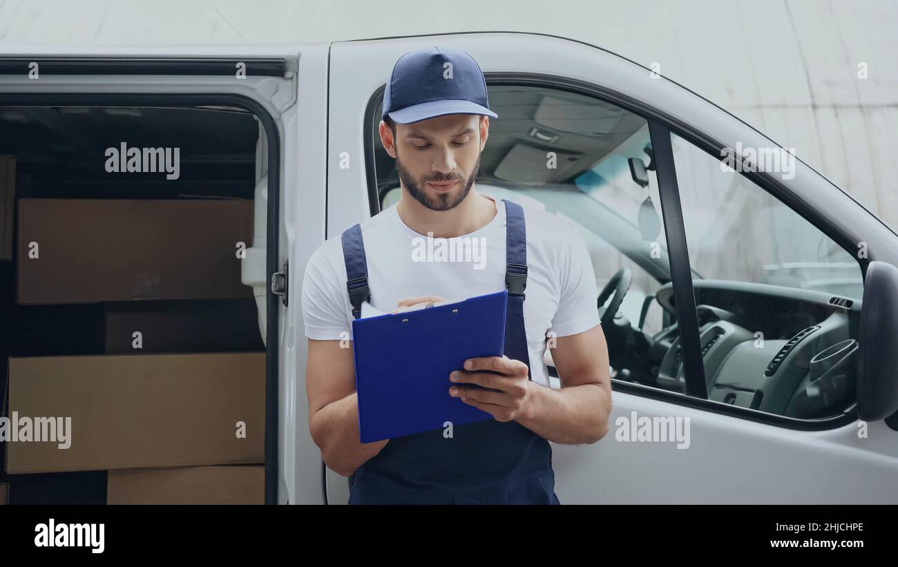 Delivery man writing on clipboard near carton boxes in car outdoors ...