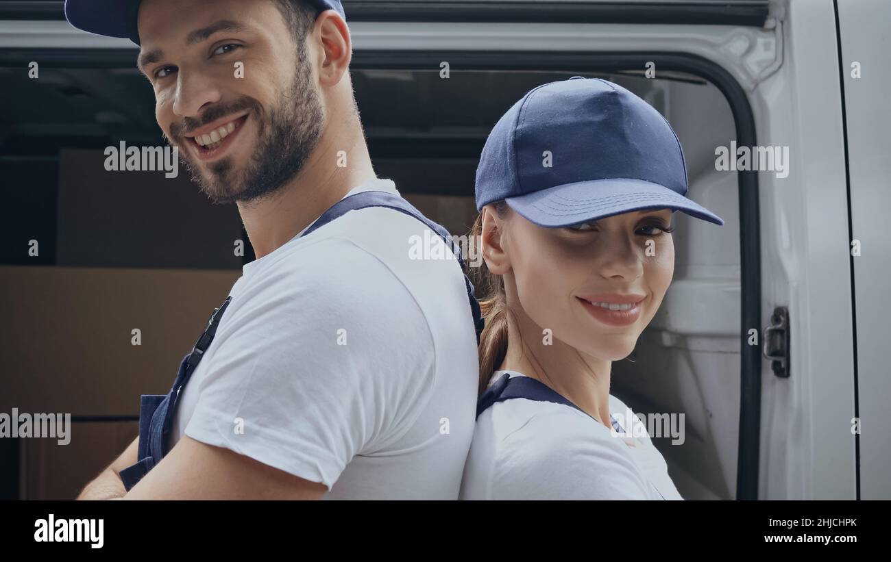 Smiling couriers looking at camera while standing back to back near car ...