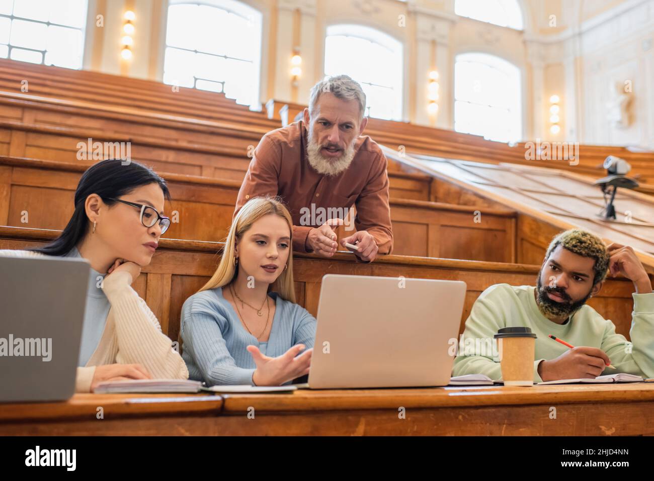 Multicultural students looking at laptop near teacher with chalk in ...