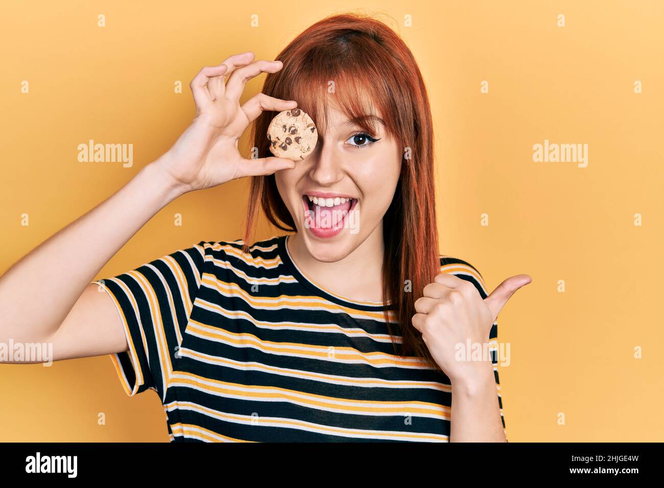 Redhead young woman holding cookie close to eye pointing thumb up to ...