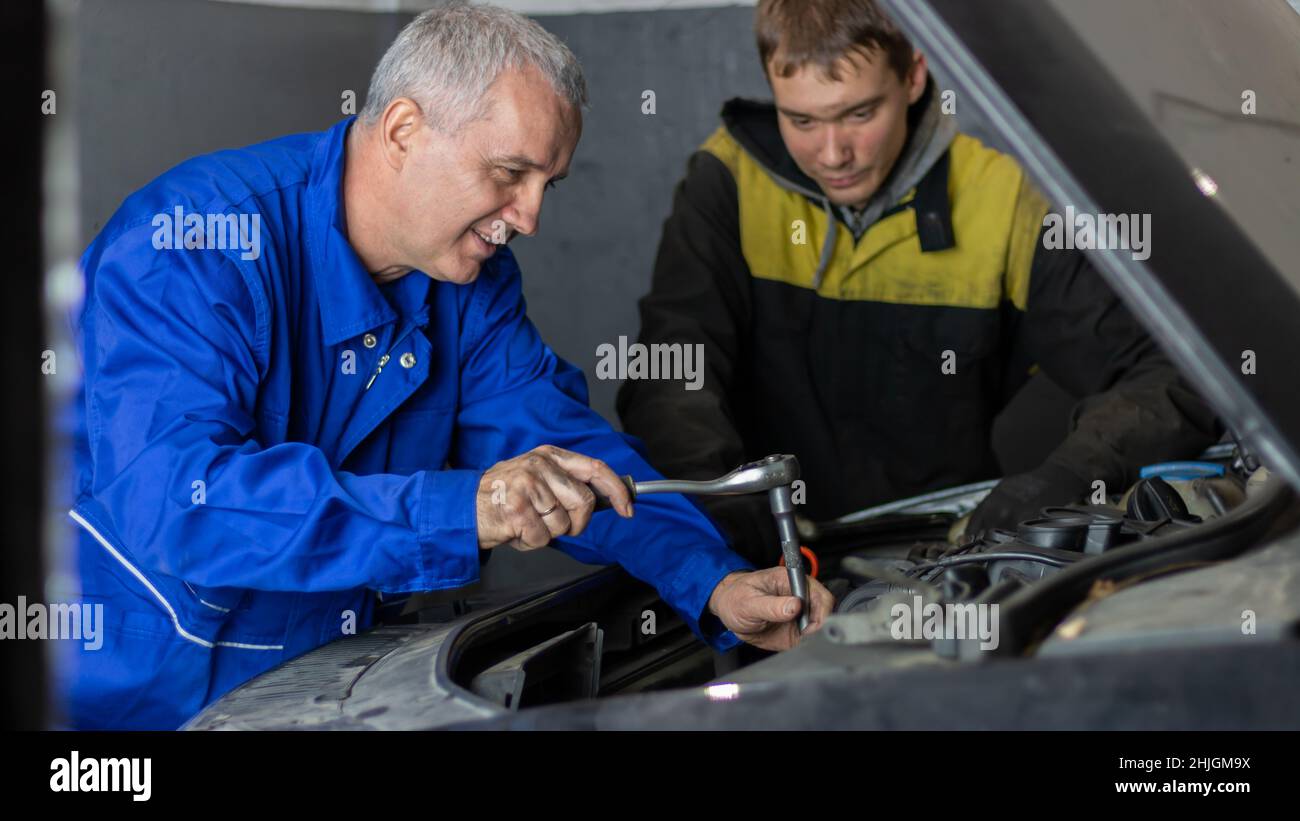 Senior experienced mechanic teaches his colleague how to repair a car ...