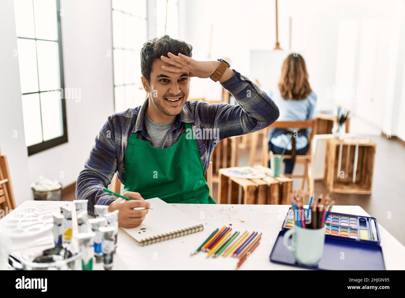 Young artist man at art studio very happy and smiling looking far away ...