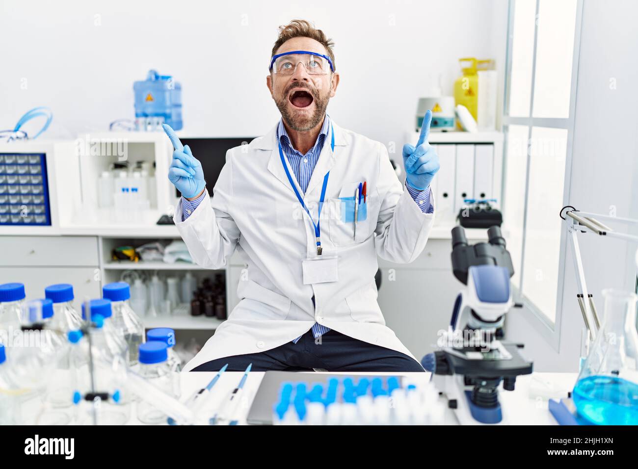 Middle age man working at scientist laboratory amazed and surprised looking up and pointing with ...