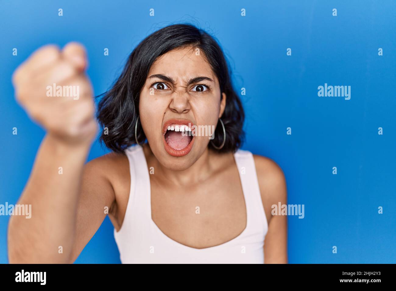 Young hispanic woman standing over blue background angry and mad ...