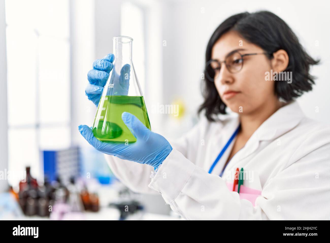 Young latin woman wearing scientist uniform measuring liquid on test ...