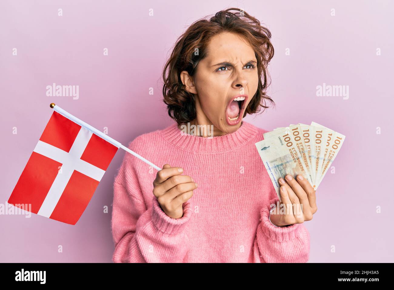 Young brunette woman holding norway flag and krone banknotes angry and ...