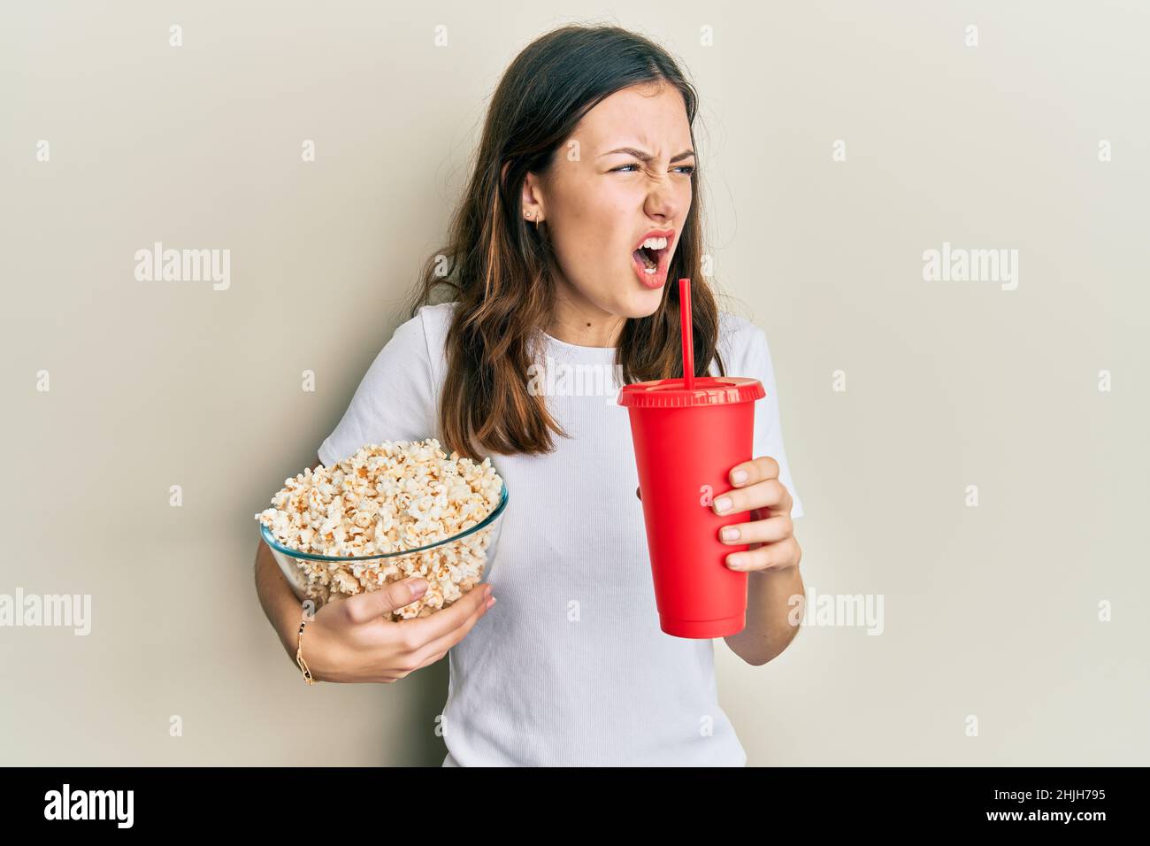Young brunette woman eating popcorn and drinking soda angry and mad ...