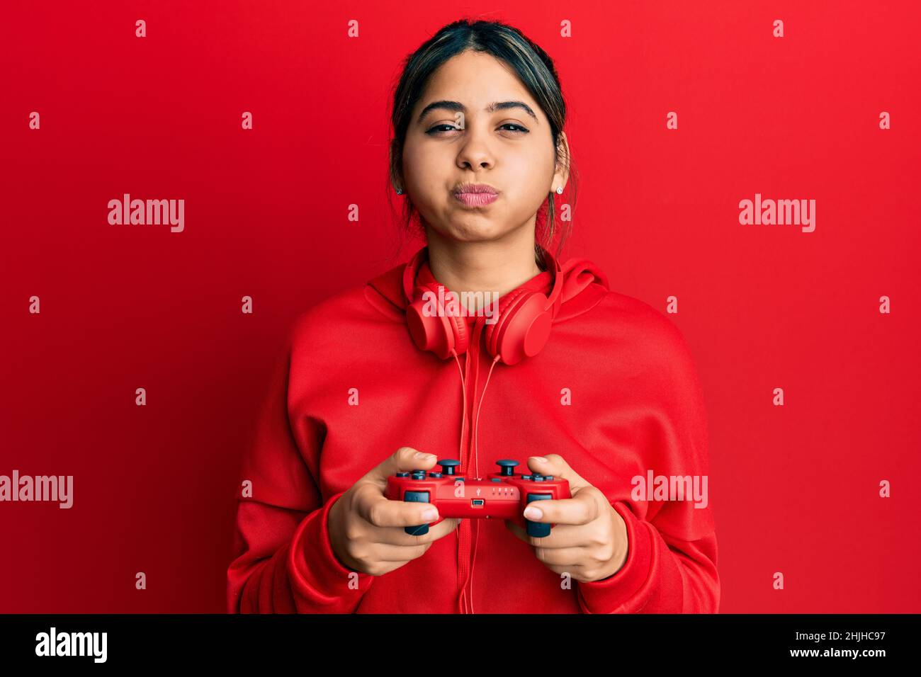 Young latin woman playing video game holding controller puffing cheeks ...