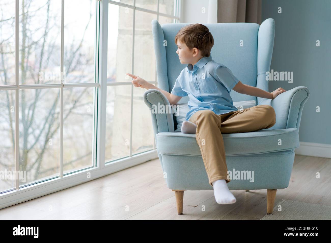 Cute boy sits in chair by window and looks out for something in window ...