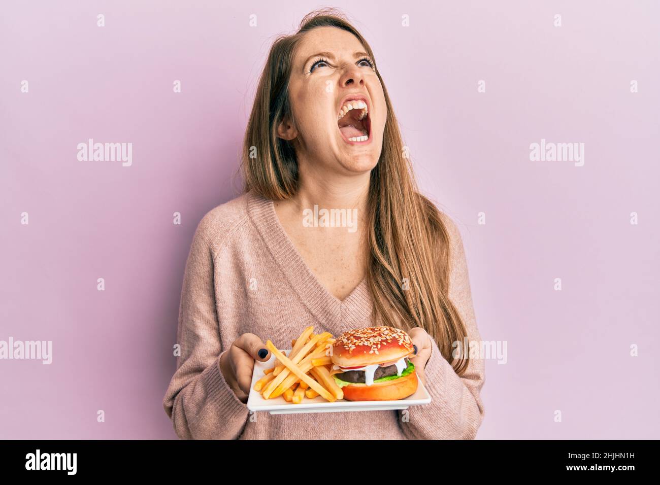 Young blonde woman eating a tasty classic burger with fries angry and ...