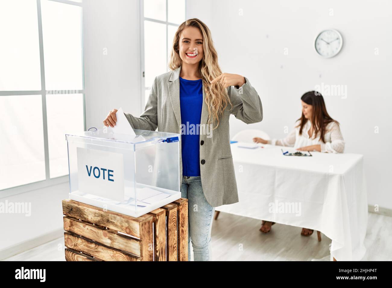 Young blonde woman voting putting envelop in ballot box pointing finger ...