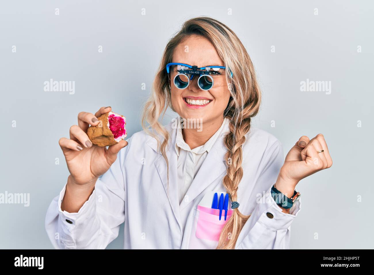 Beautiful young blonde woman holding geode stone wearing magnifier ...