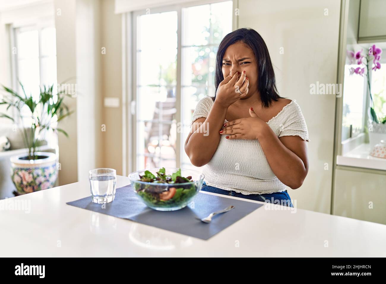 Young hispanic woman eating healthy salad at home smelling something ...