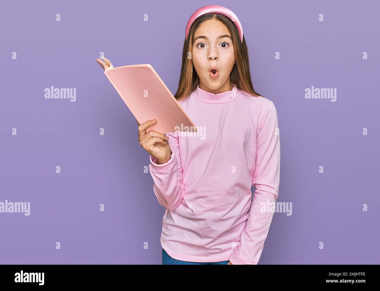 Beautiful brunette little girl reading a book scared and amazed with ...