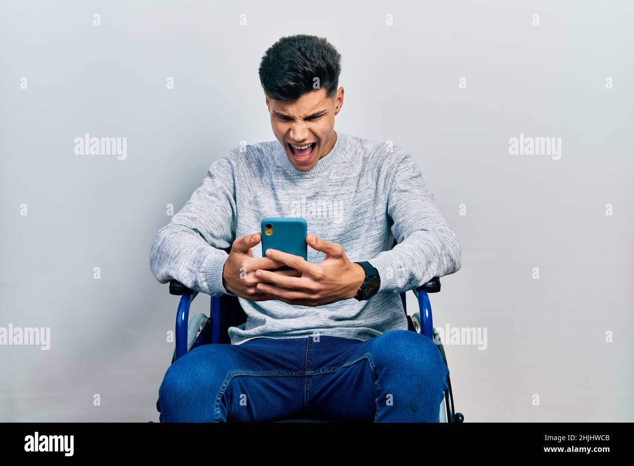 Young hispanic man sitting on wheelchair using smartphone angry and mad ...