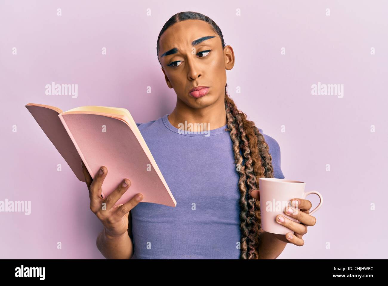 Hispanic man wearing make up and long hair reading book drinking cup of ...