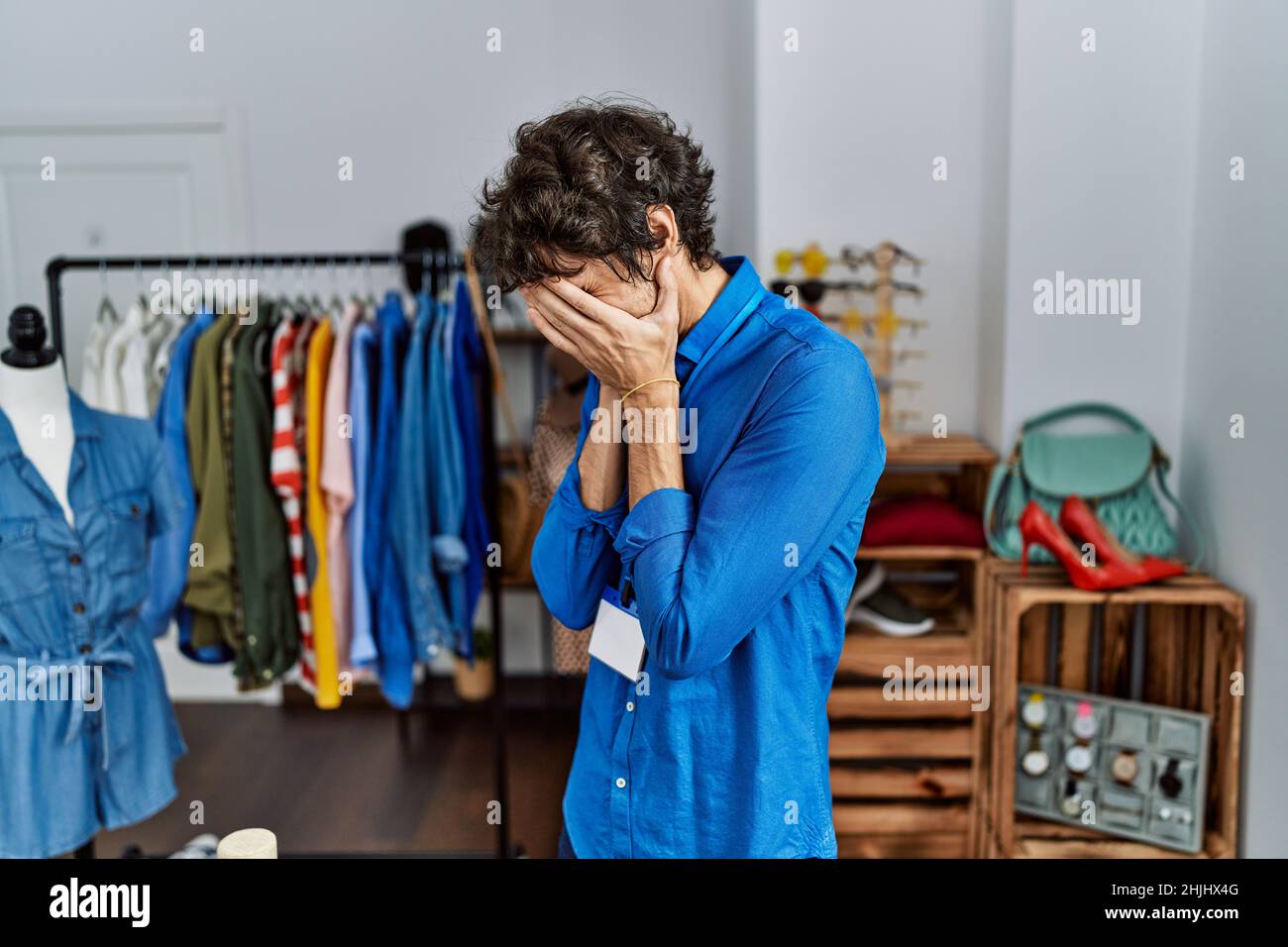 Young hispanic man working as manager at retail boutique with sad expression covering face with ...