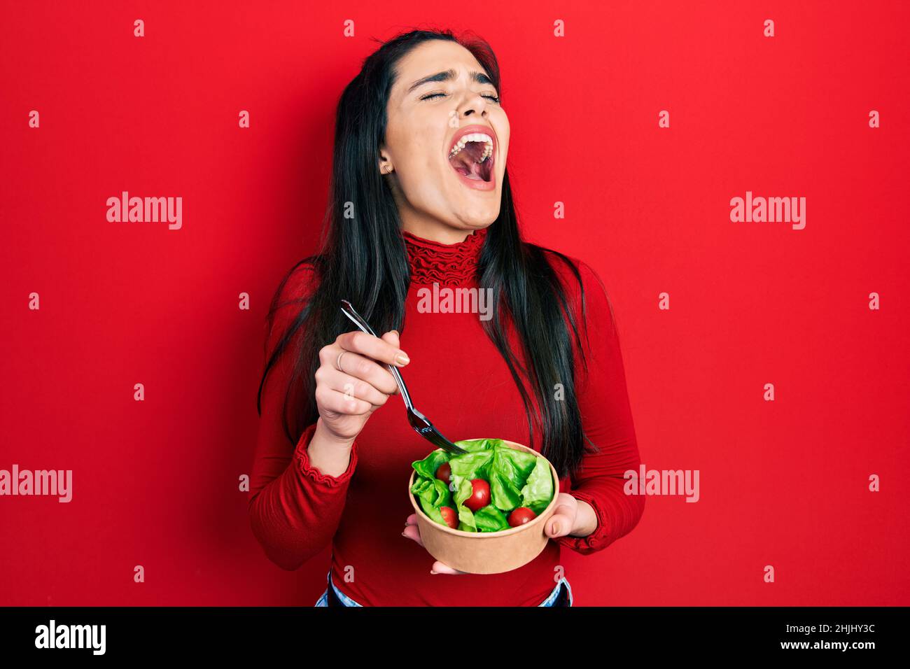 Young hispanic girl eating salad angry and mad screaming frustrated and ...