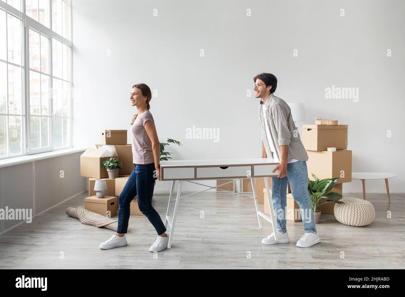 Cheerful european millennial man and lady carry table in empty room ...