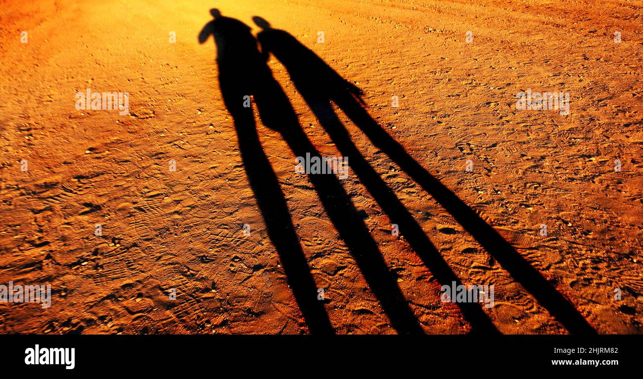 Long shadows of two people holding hands walking down a dirt road ...