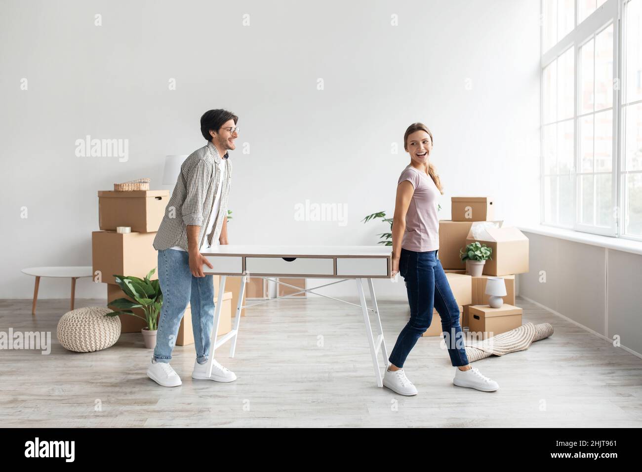 Cheerful millennial caucasian wife and husband carry table in room ...