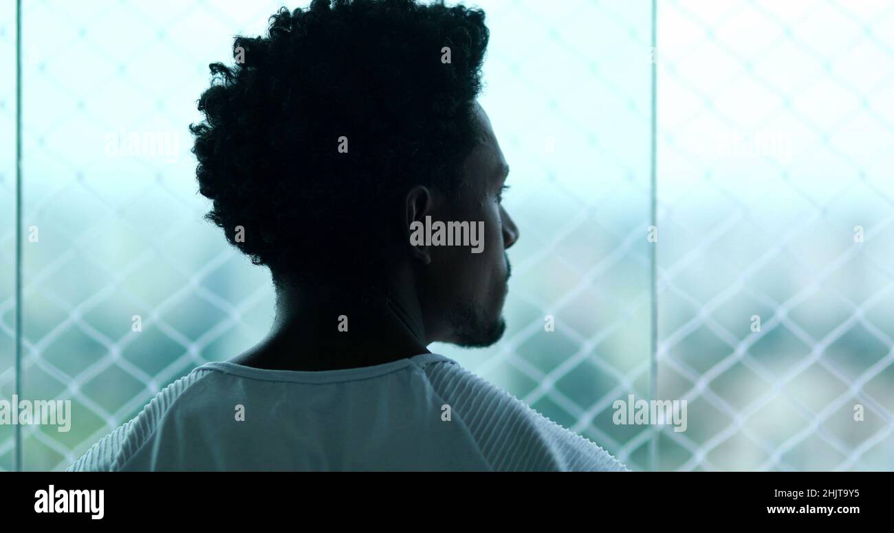 Person standing by apartment window looking outside. Pensive black man ...