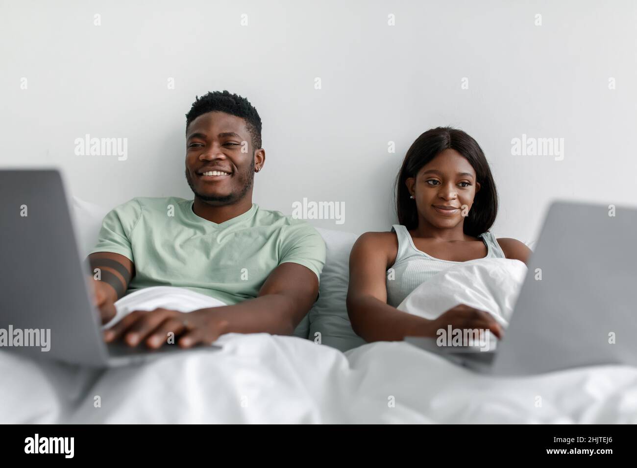 Happy millennial african american wife and husband lie on bed with computers, play games Stock ...