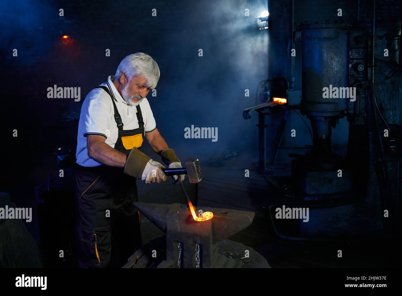 Aged caucasian blacksmith wearing safety apron and gloves forging steel ...