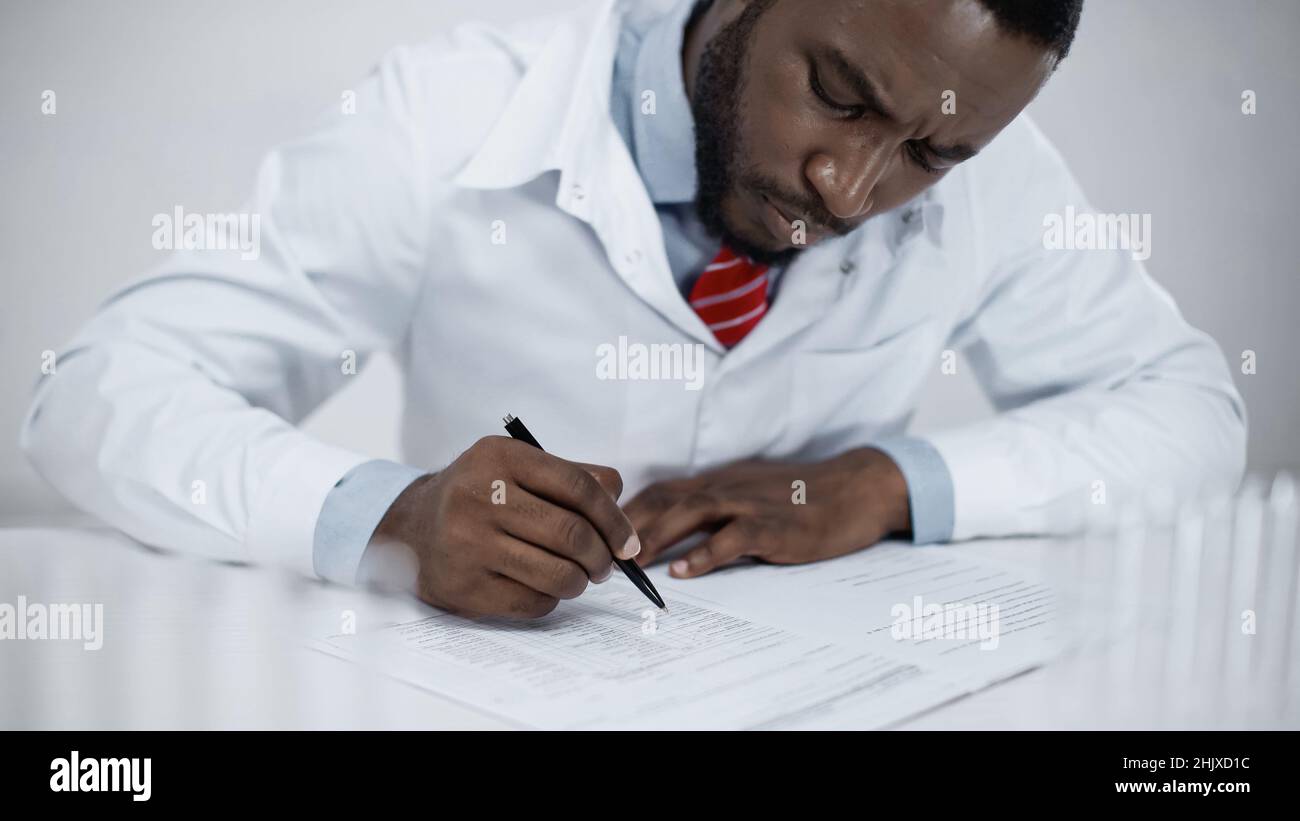 focused african american doctor in white coat signing documents in ...