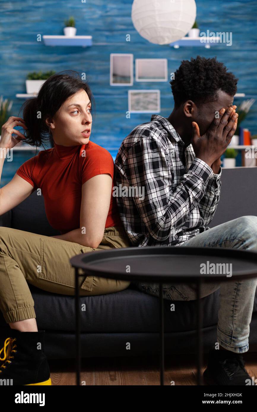 Displeased interracial couple sitting on sofa in silence while ...