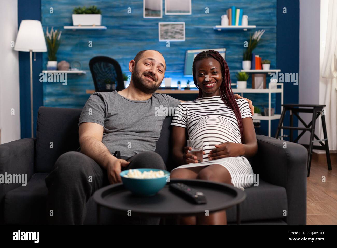 Multiracial couple enjoying television and expecting baby at home while ...