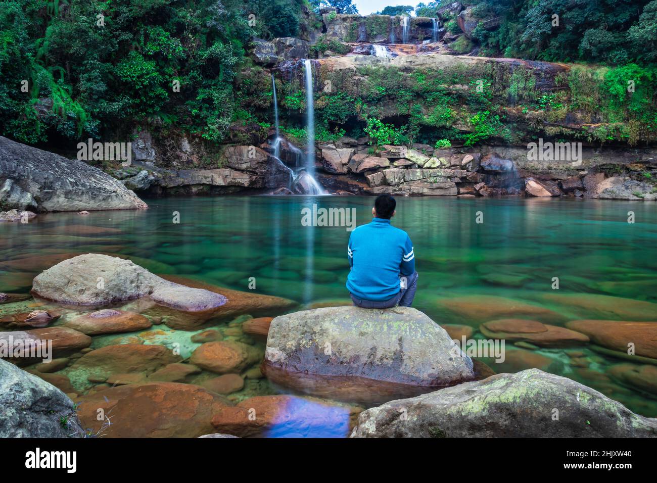 young man isolated sitting on rock with pristine waterfall background ...
