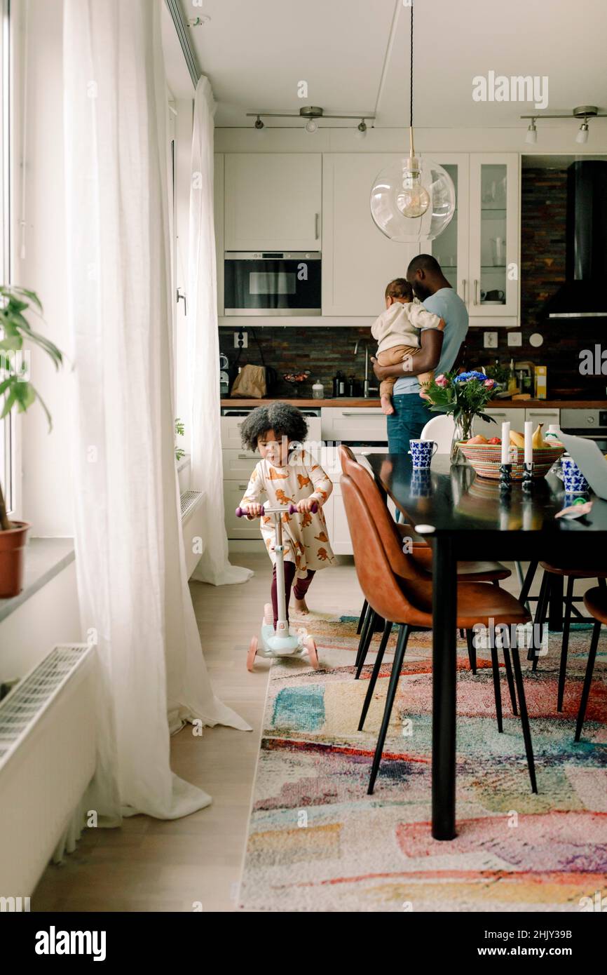 Father carrying baby boy while daughter riding push scooter in kitchen ...