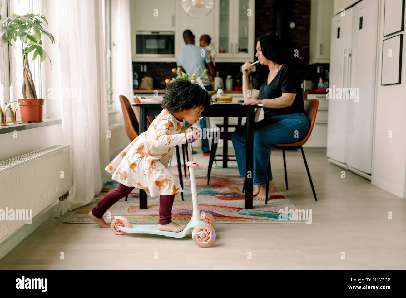 Daughter riding push scooter while mother working in kitchen Stock ...