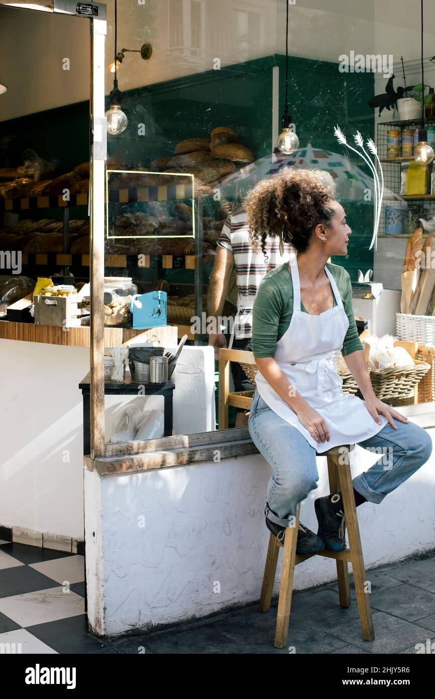 Female owner looking away while sitting on stool outside bakery Stock ...