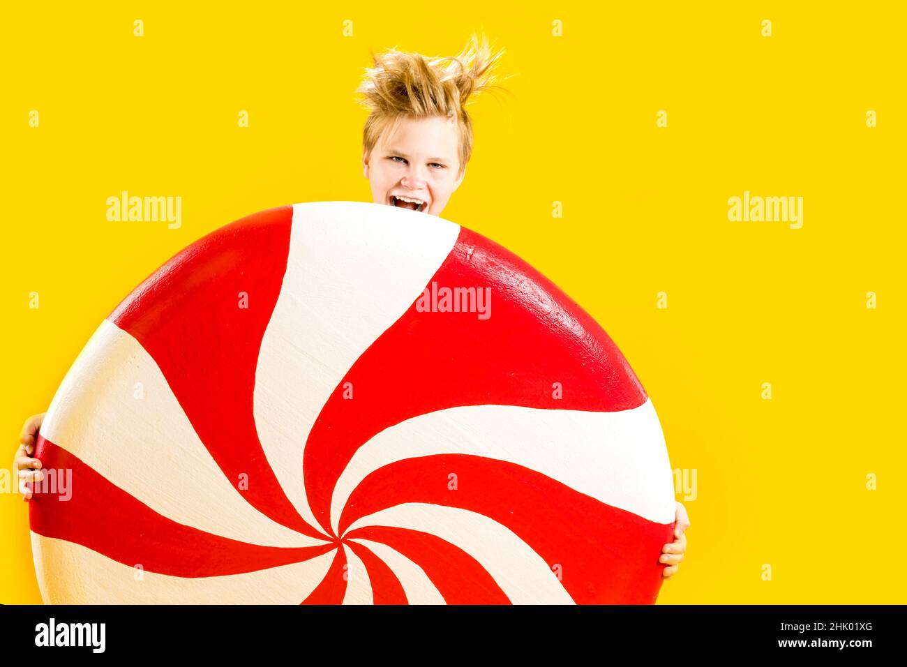A cute boy with a huge candy on a yellow background, trying to eat it ...