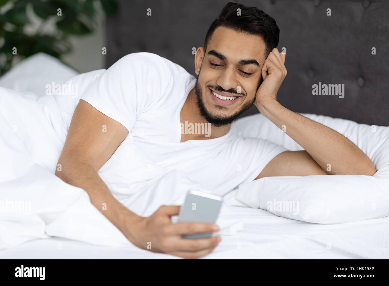 Cheerful Young Middle Eastern Guy Lying In Bed And Using Smartphone ...